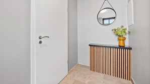 Um corredor com porta branca, um banco de ripas de madeira, um espelho redondo e um vaso com planta.