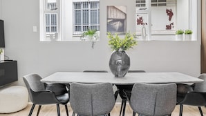 Uma área de refeições moderna com uma mesa retangular, seis cadeiras cinzas estofadas, um vaso grande com plantas verdes e uma janela grande com vidro dividido.