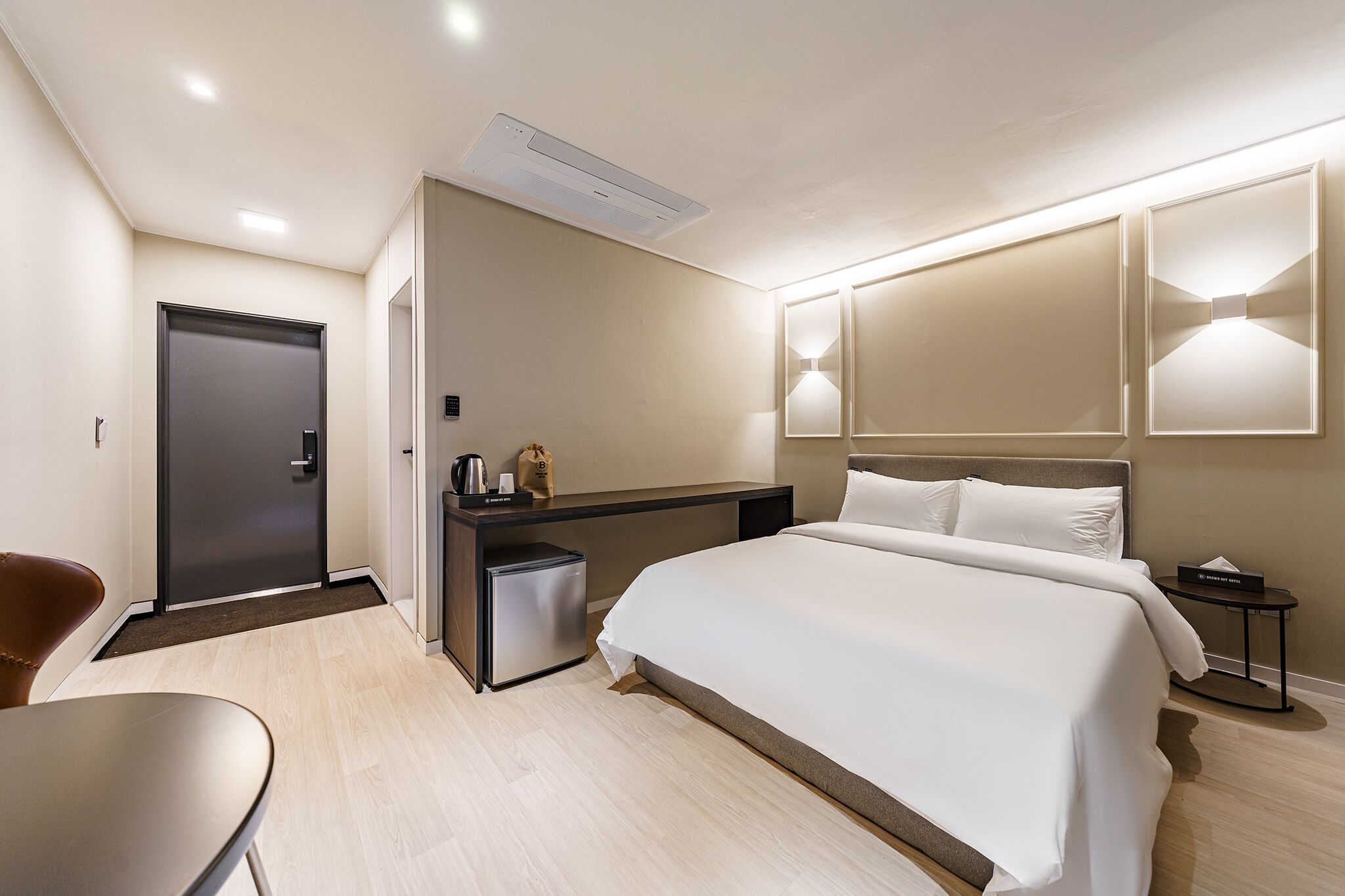 Quarto de hotel moderno com uma cama grande, uma escrivaninha, uma cadeira e uma mesinha de cabeceira com abajur.