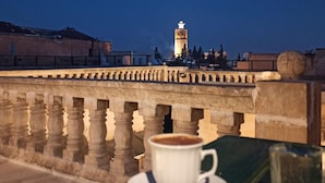 Varanda com uma xícara de café e uma tigela de fruta, com vista para uma torre e paisagem urbana ao entardecer.