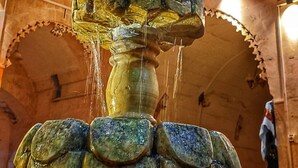 Uma fonte de pedra em vários níveis, com água a correr do nível superior, em frente a uma fachada arquitetônica tradicional com arcos e varanda.