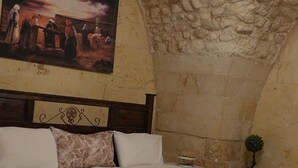 Quarto com teto de pedra, cama com lençóis brancos, um quadro na parede e uma pequena planta em vaso.