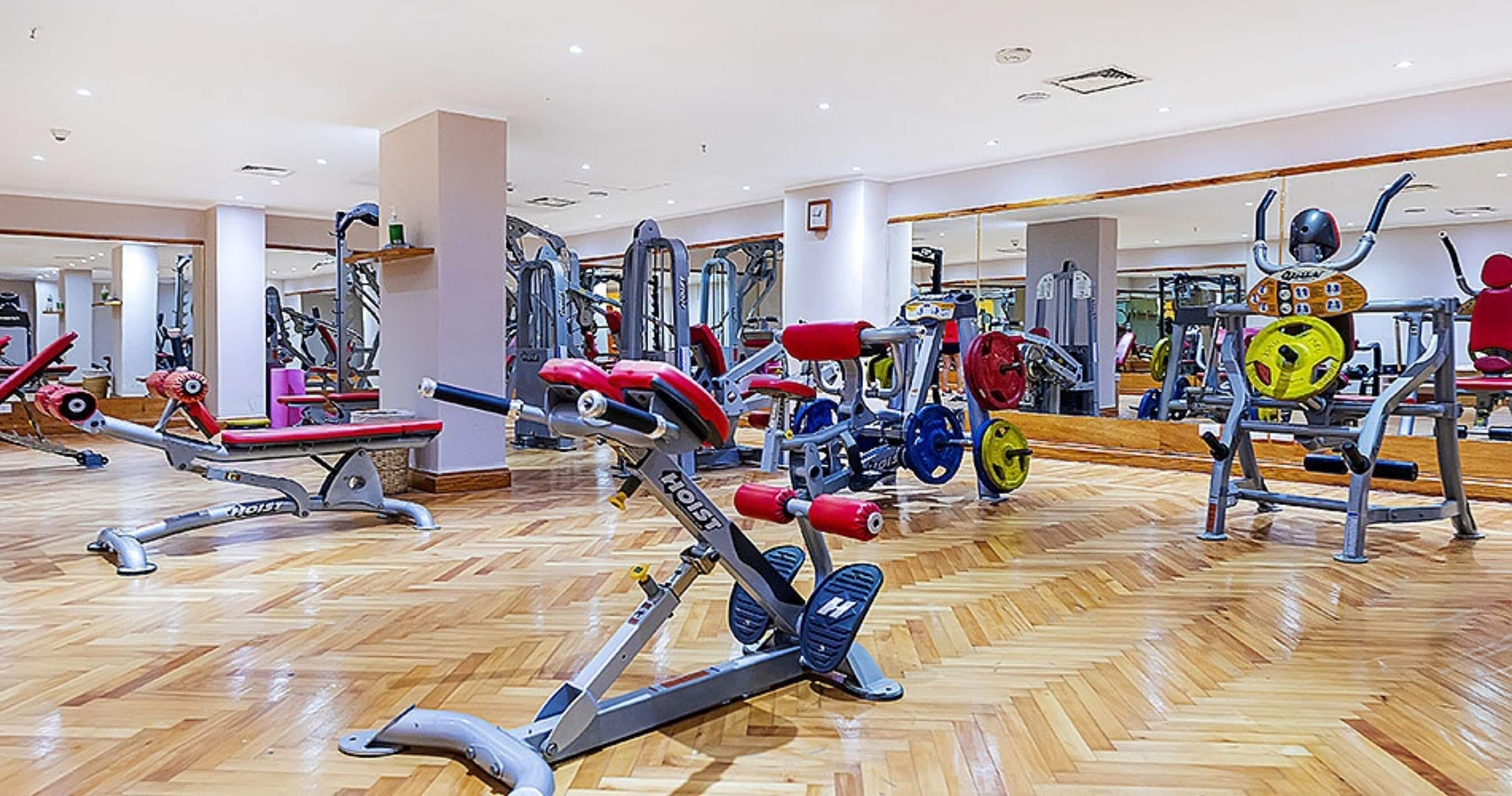 Uma academia com diversos equipamentos de exercícios, piso de madeira e espelhos nas paredes.
