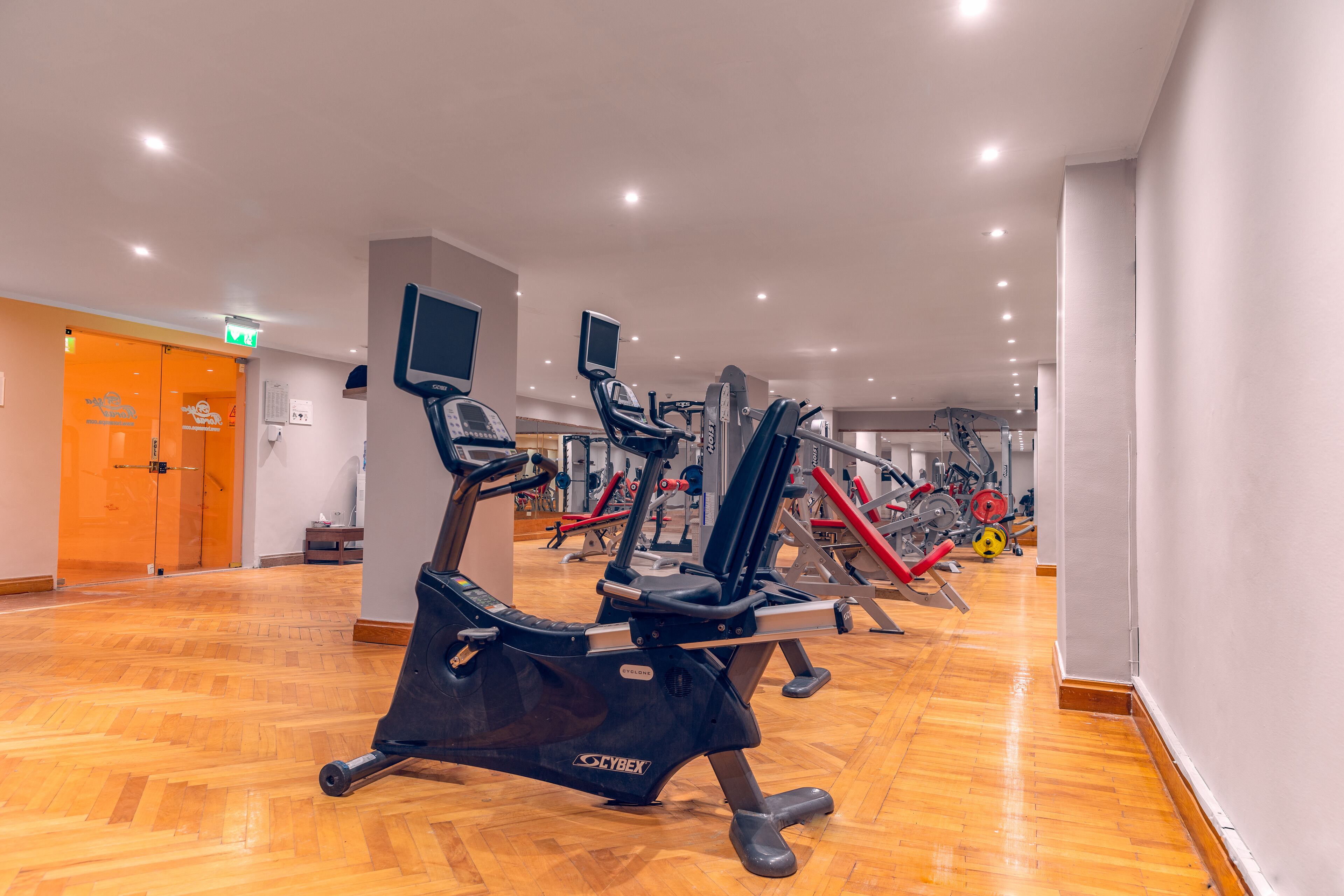 Uma academia com máquinas de cardio, equipamentos de musculação e piso de madeira.
