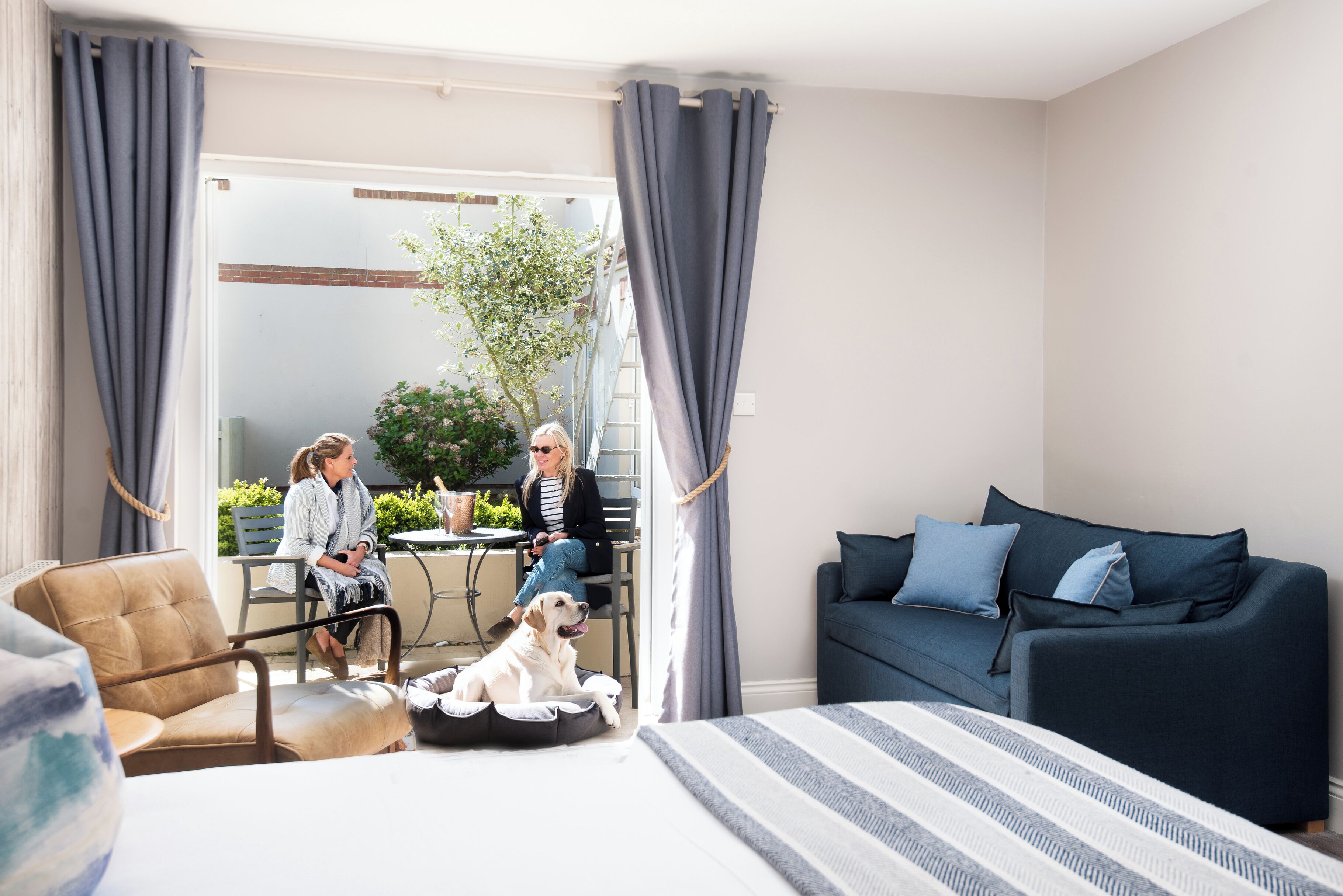 Quarto de hotel com cama, sofá, caminha para cachorro, cadeira e mesa com duas pessoas e um cão.