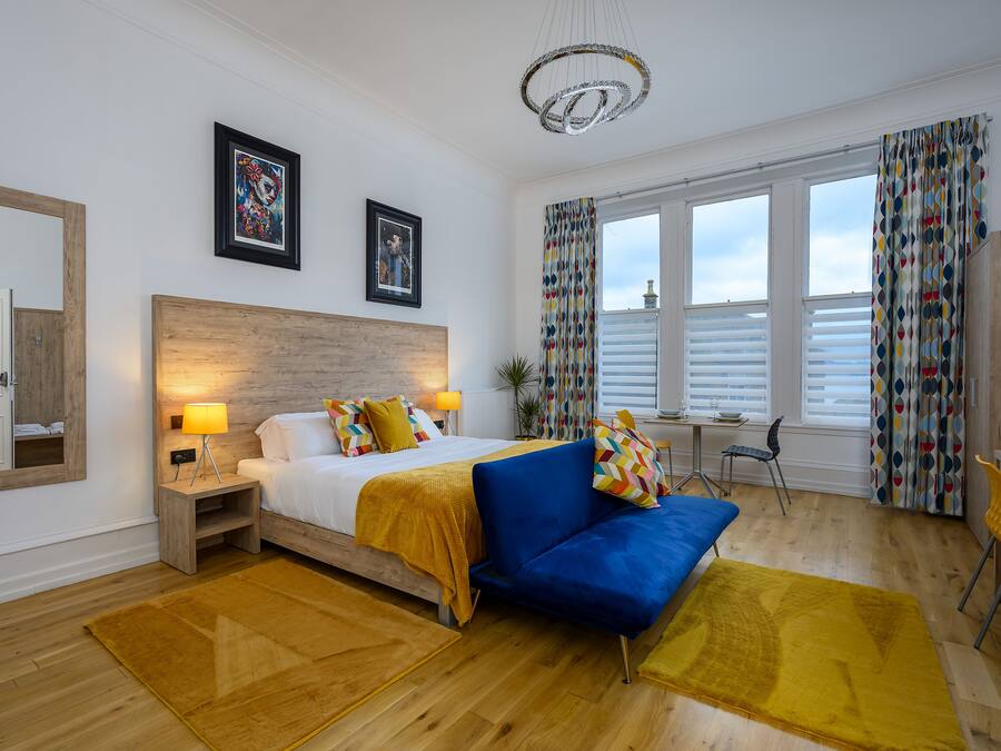 Quarto moderno com cama e cabeceira de madeira, um sofá azul e um tapete amarelo. Há duas luminárias de mesa, um espelho e uma janela com cortinas.