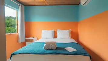 Quarto de hotel com cama, mesa de cabeceira, janela e vista para vegetação, e aparelho de ar condicionado.