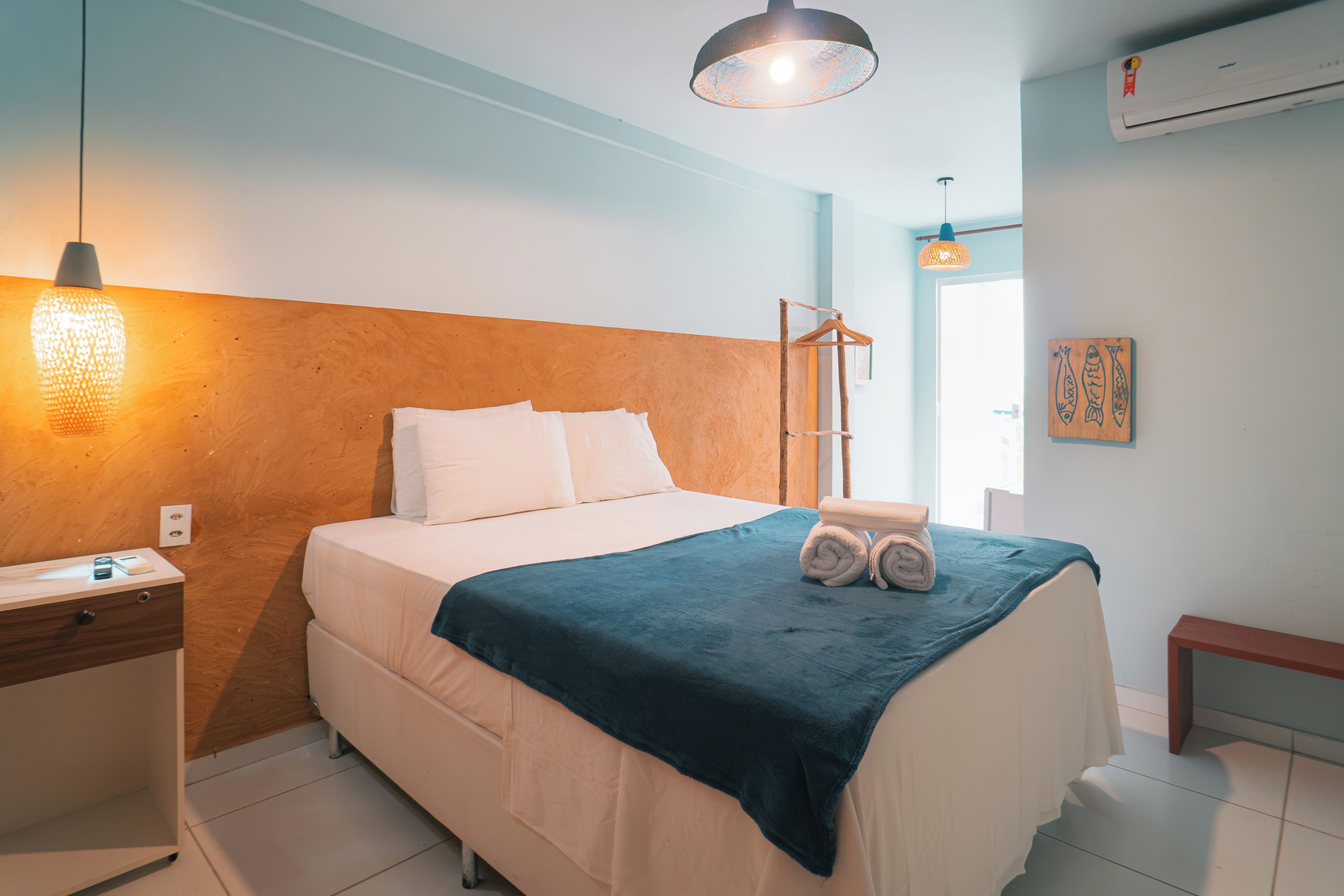 Quarto com cama, mesa de cabeceira, luminária pendente e ar-condicionado fixado na parede.