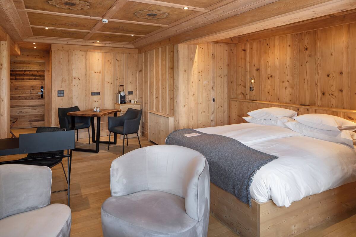 Quarto de madeira com uma cama, duas cadeiras, uma mesa e um relógio na parede.