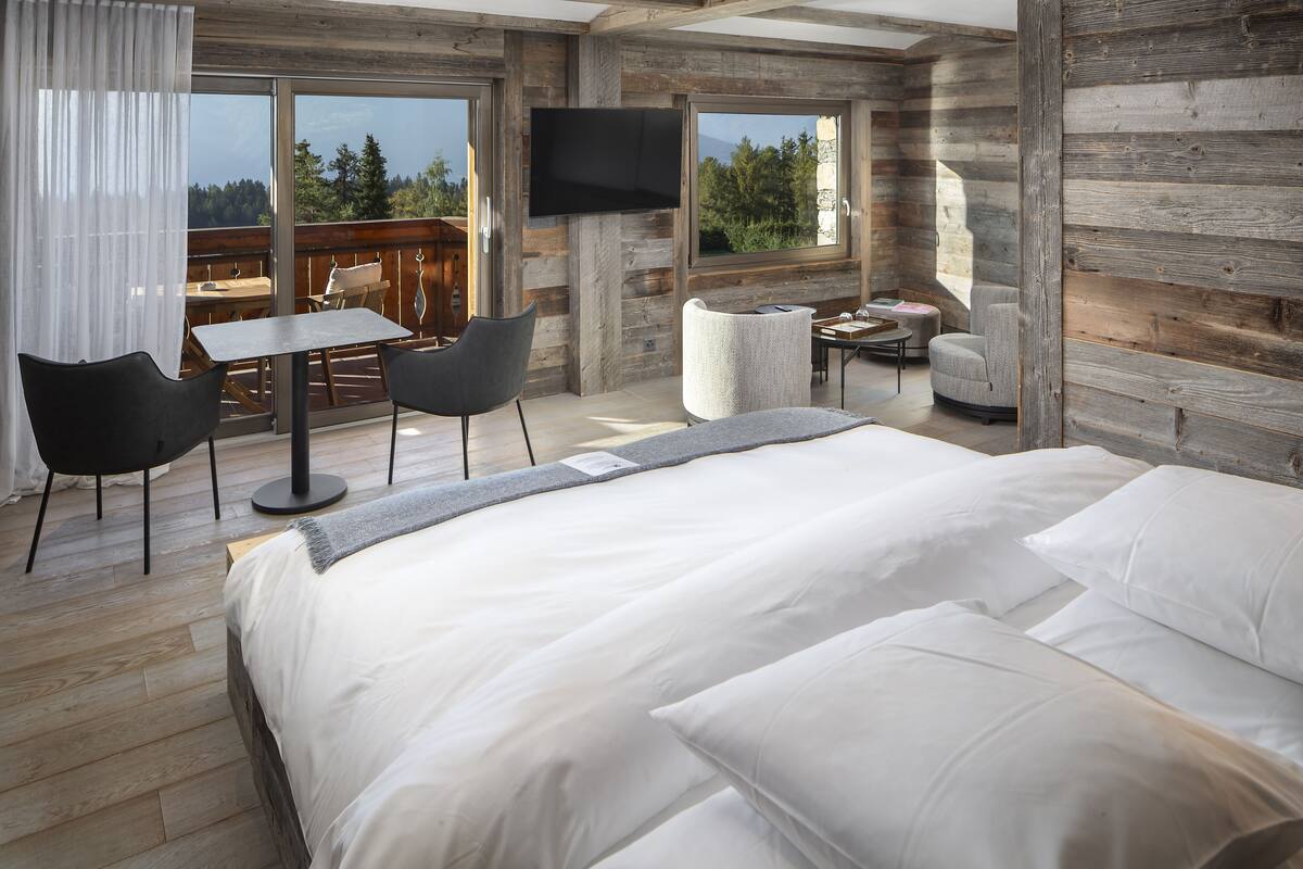 Quarto com cama grande, televisão, mesa de jantar com cadeiras e varanda com vista para árvores.
