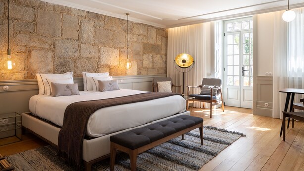Quarto com uma cama grande, um banco, uma cadeira, uma escrivaninha e uma parede de pedra.