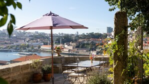 Um pátio externo com mesa, cadeiras, um guarda-sol roxo e vasos de plantas.