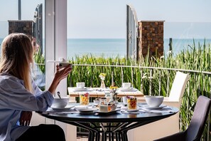 Uma pessoa à mesa com vista para o mar, varanda com grades e uma mesa posta com pratos e um copo de fruta.