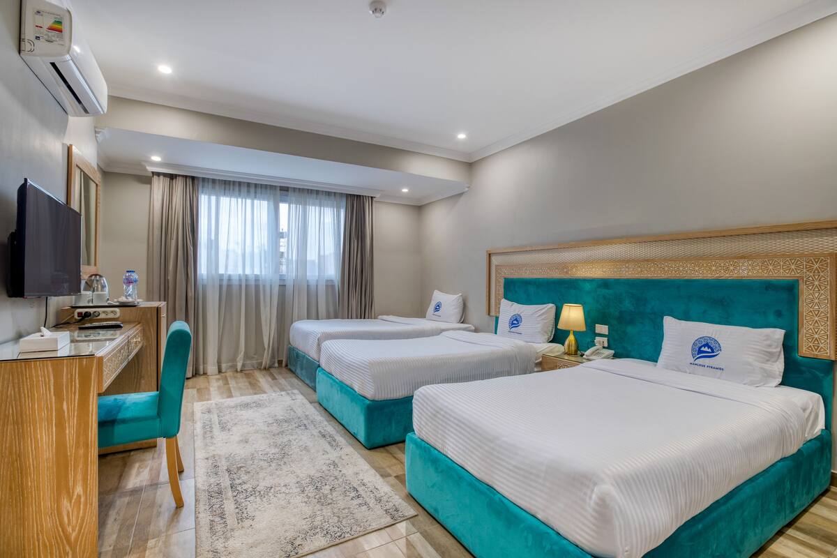 Quarto de hotel com duas camas, uma escrivaninha, uma cadeira, uma televisão e uma janela com cortinas.