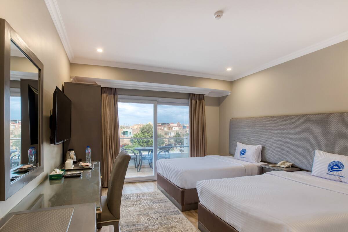 Quarto de hotel com duas camas, uma escrivaninha com cadeira, televisão e varanda com vista para edifícios.