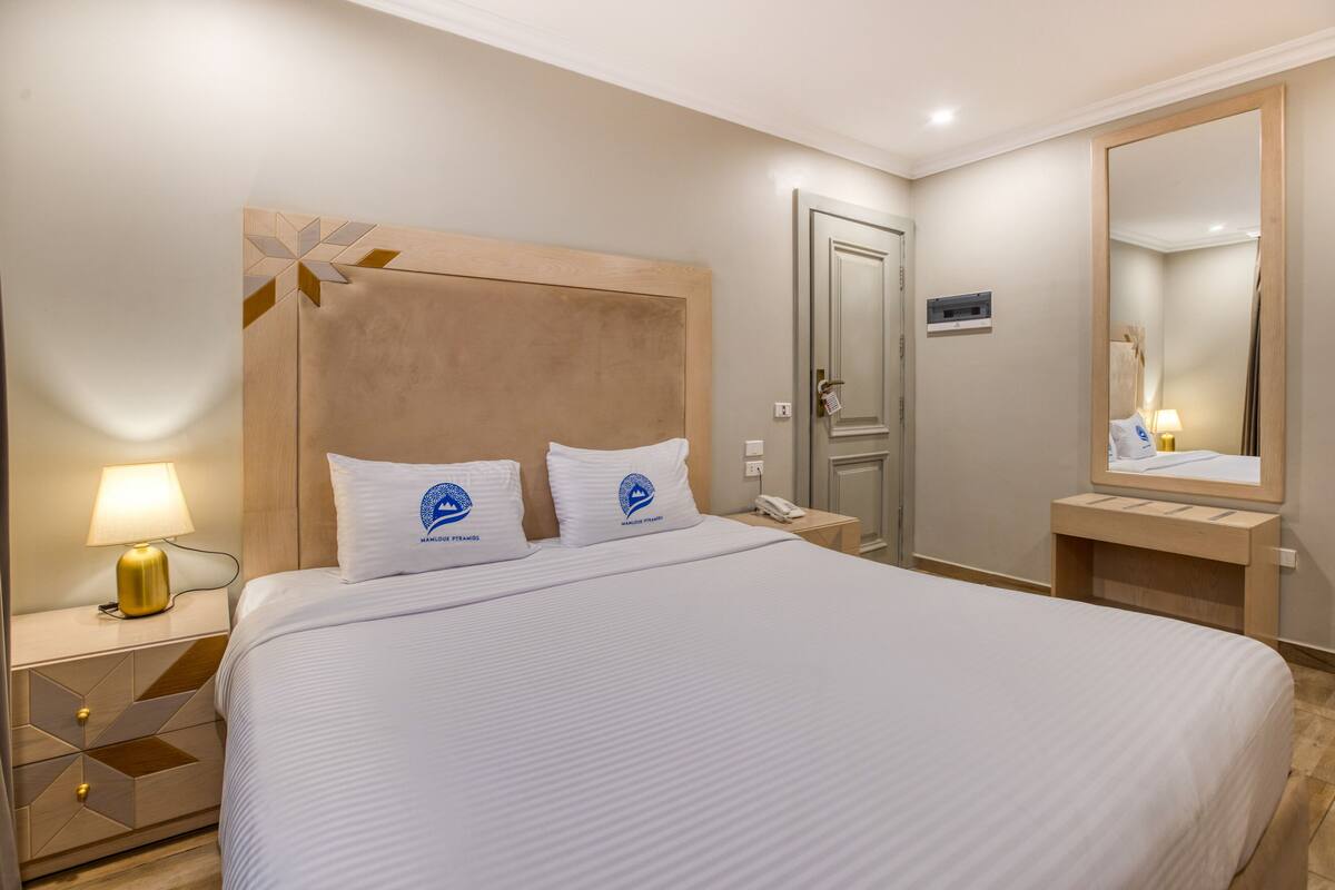 Quarto de hotel com uma cama grande, duas almofadas com logotipo, abajur, mesa de cabeceira, espelho e uma porta.