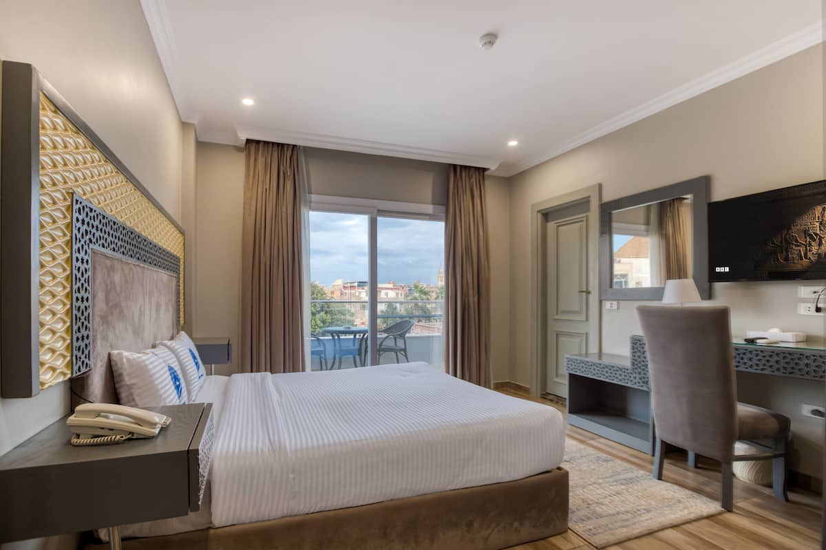 Quarto de hotel com uma cama grande, uma mesa com cadeira, televisão e varanda com vista para edifícios.