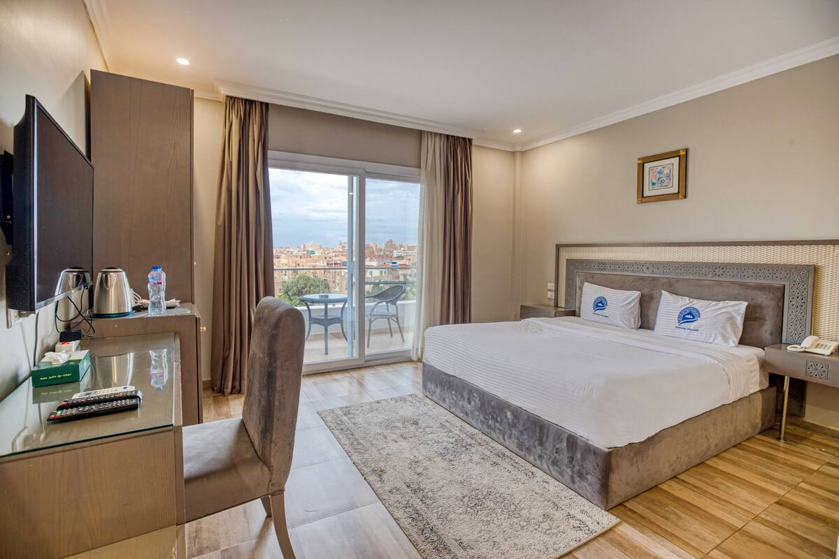 Quarto de hotel com uma cama grande, uma escrivaninha com uma TV, uma cadeira e vista para a cidade pela janela.