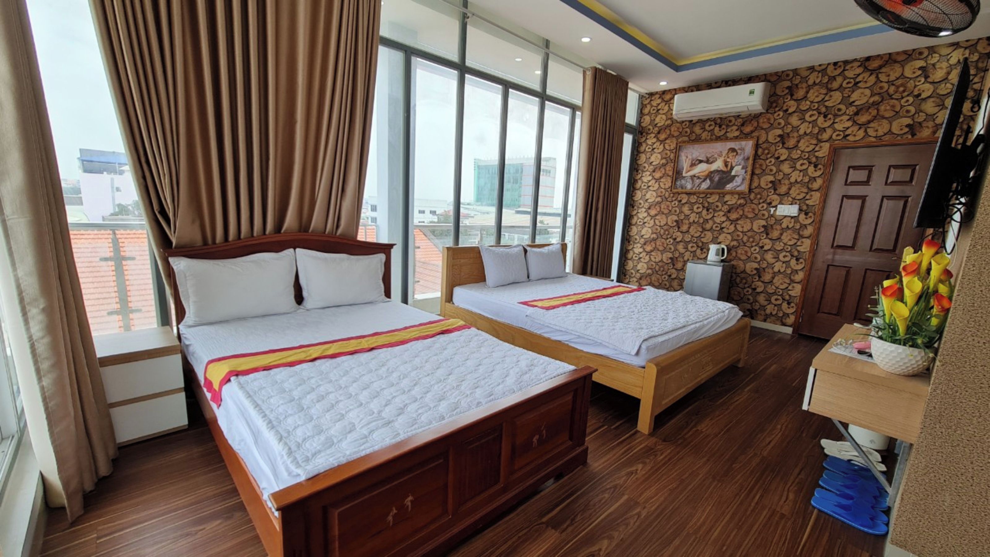 Quarto de hotel com duas camas de madeira, amplas janelas, vista para a cidade e um detalhe decorativo na parede.