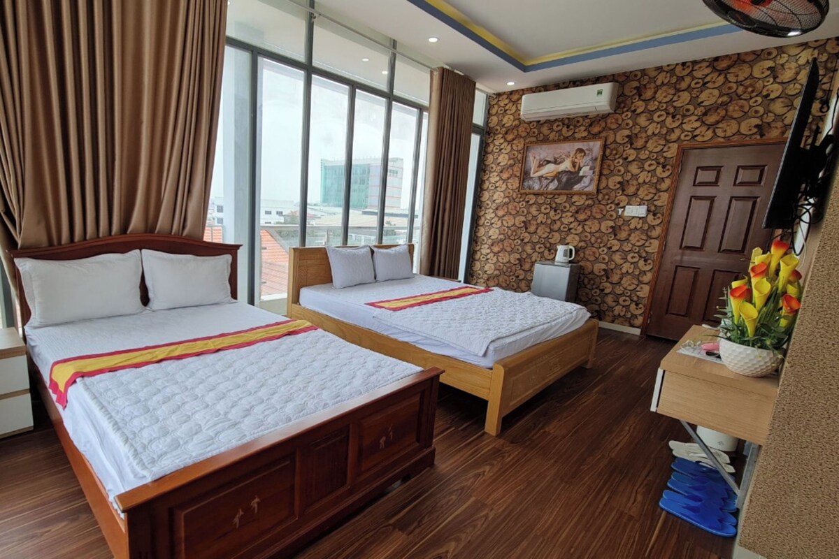 Quarto de hotel com duas camas de madeira, amplas janelas, vista para a cidade e um detalhe decorativo na parede.