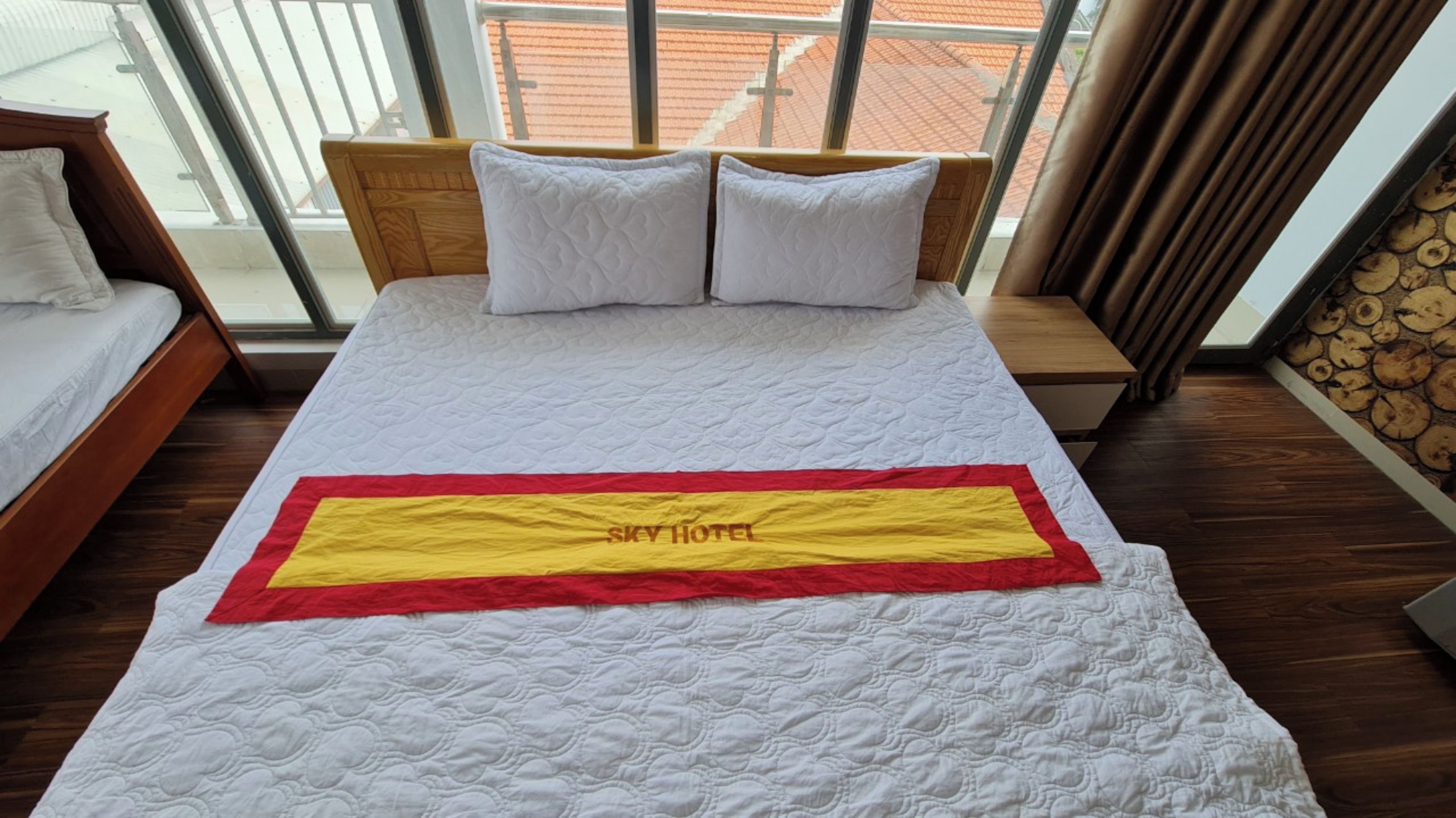 Quarto de hotel com cama, travesseiros, cabeceira de madeira, janela com vista e colcha vermelha e amarela com o texto 'SKY HOTEL'.