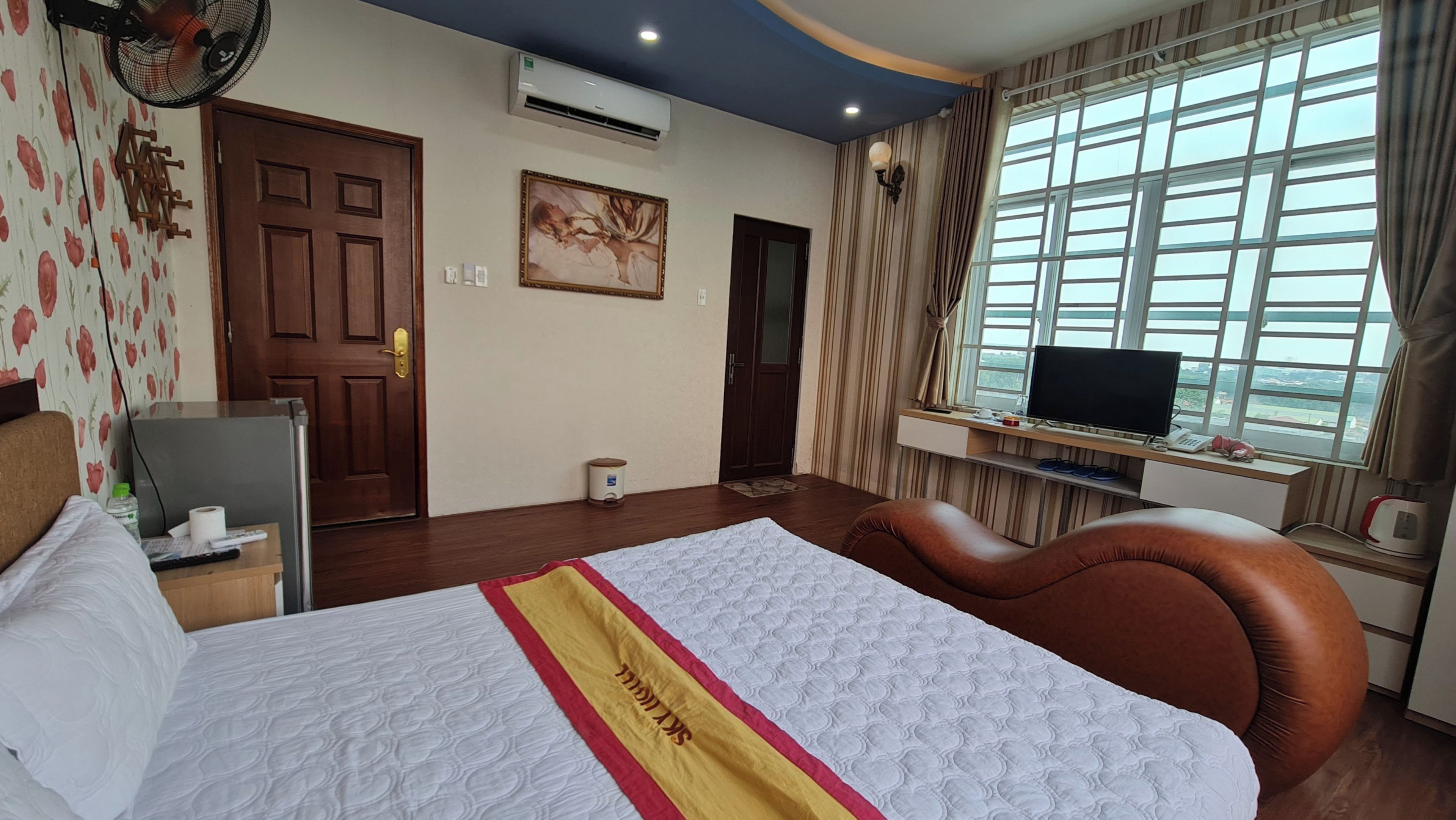 Quarto de hotel com cama, escrivaninha com computador, televisão e janela com cortinas.