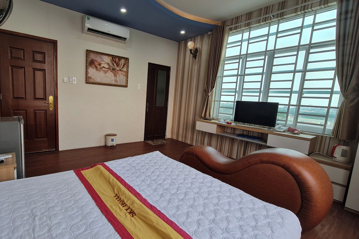 Quarto de hotel com cama, escrivaninha com computador, televisão e janela com cortinas.