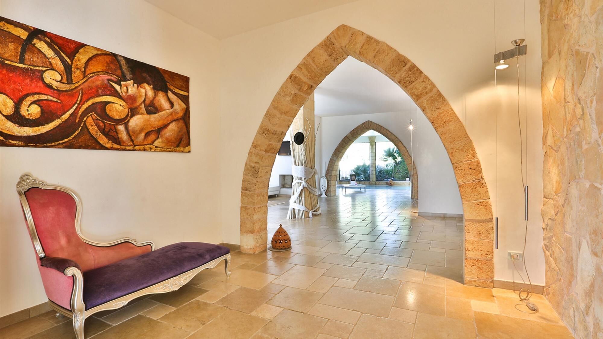 Um hall com uma poltrona de veludo vermelho, uma grande pintura abstrata e uma porta em arco que dá para uma área ampla com piso de pedra e um lustre.