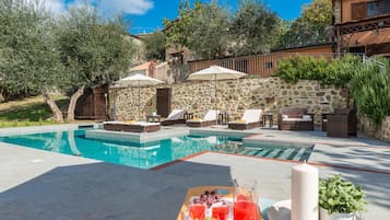 Piscina com espreguiçadeiras e guarda-sóis, cercada por muros de pedra e vegetação.
