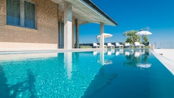Piscina com água azul cristalina, rodeada por um edifício de tijolo com grandes janelas. Há espreguiçadeiras e guarda-sóis ao longo da piscina.
