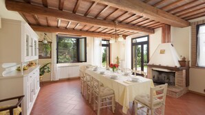 Uma sala de jantar com uma mesa comprida posta para seis, vigas de madeira no teto, lareira e vista para o jardim através de grandes janelas.