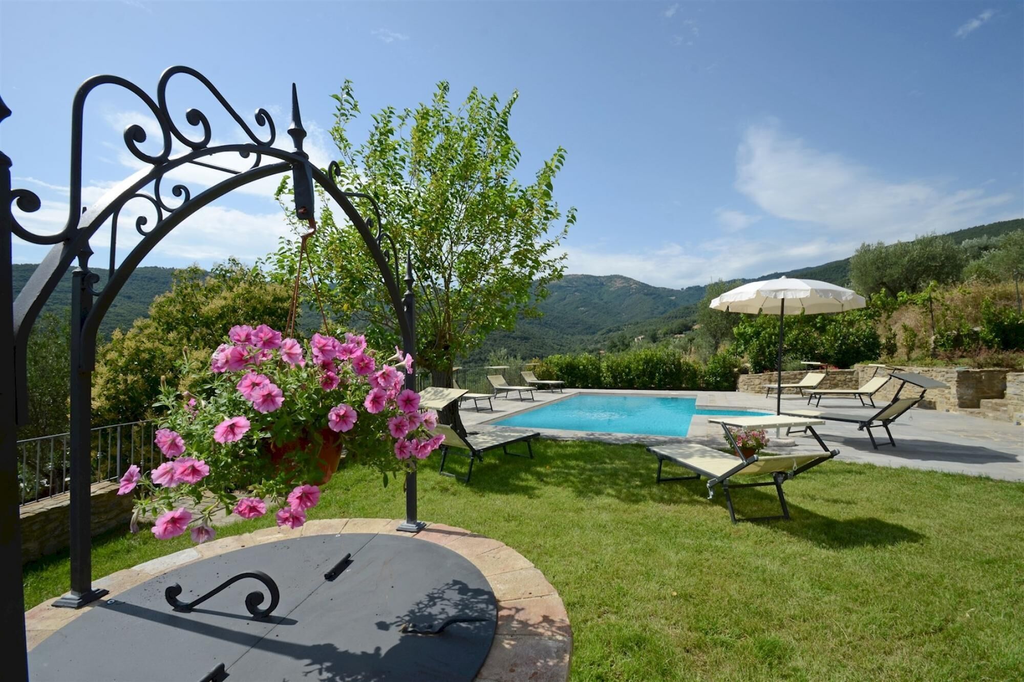 Uma área na piscina com vista para montanhas, um pérgola com flores cor-de-rosa e espreguiçadeiras.
