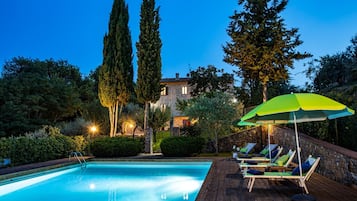 Uma piscina com espreguiçadeiras e um guarda-sol, rodeada por árvores e um edifício de pedra.