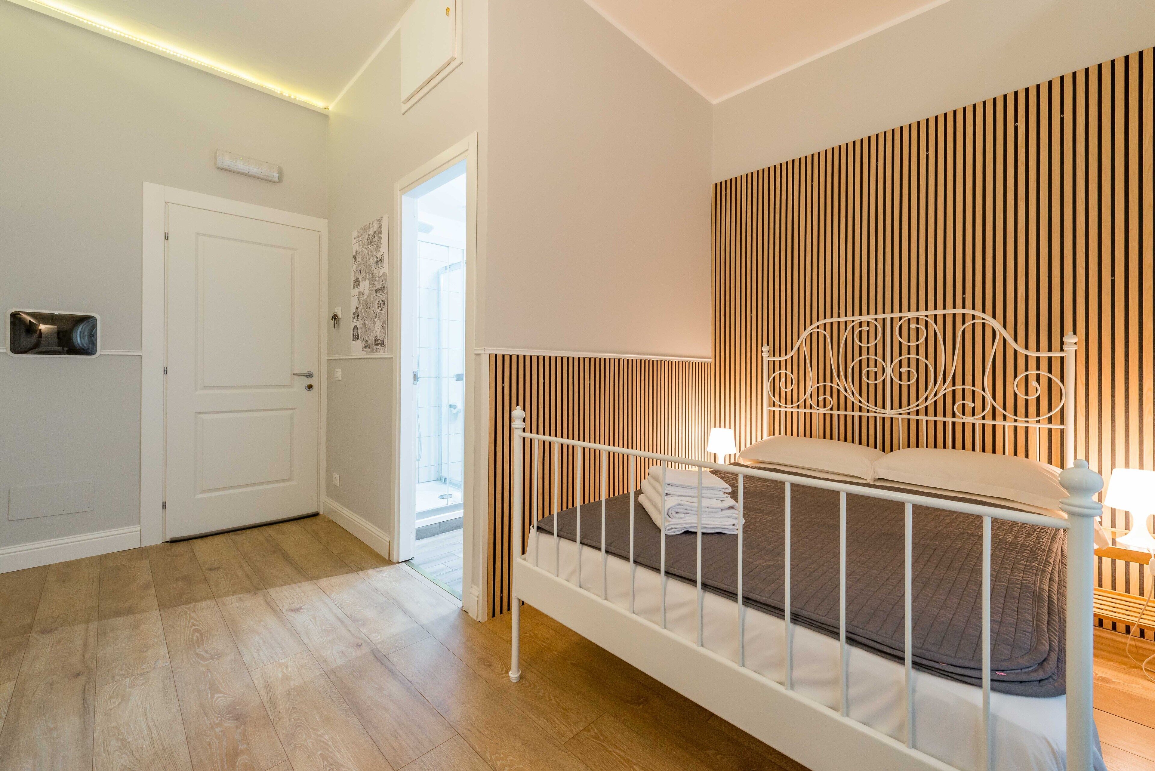 Quarto com estrutura de cama de metal branco, piso de madeira, parede com ripas de madeira e porta branca.
