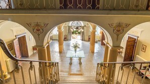 Uma escadaria imponente leva a um espaço central com uma mesa de vidro e arranjo floral, ladeado por colunas e arcos elaborados.