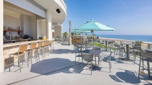 Um bar no terraço com banquetas, mesas e guarda-sóis, com vista para o oceano.