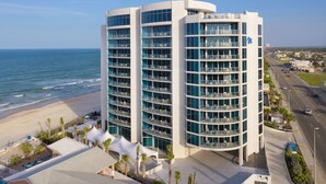 Um hotel com vários andares e varandas com vista para a praia, com uma visão clara do oceano e um estacionamento em primeiro plano.