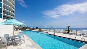 Uma piscina com espreguiçadeiras e guarda-sóis, céu claro e um edifício ao fundo.