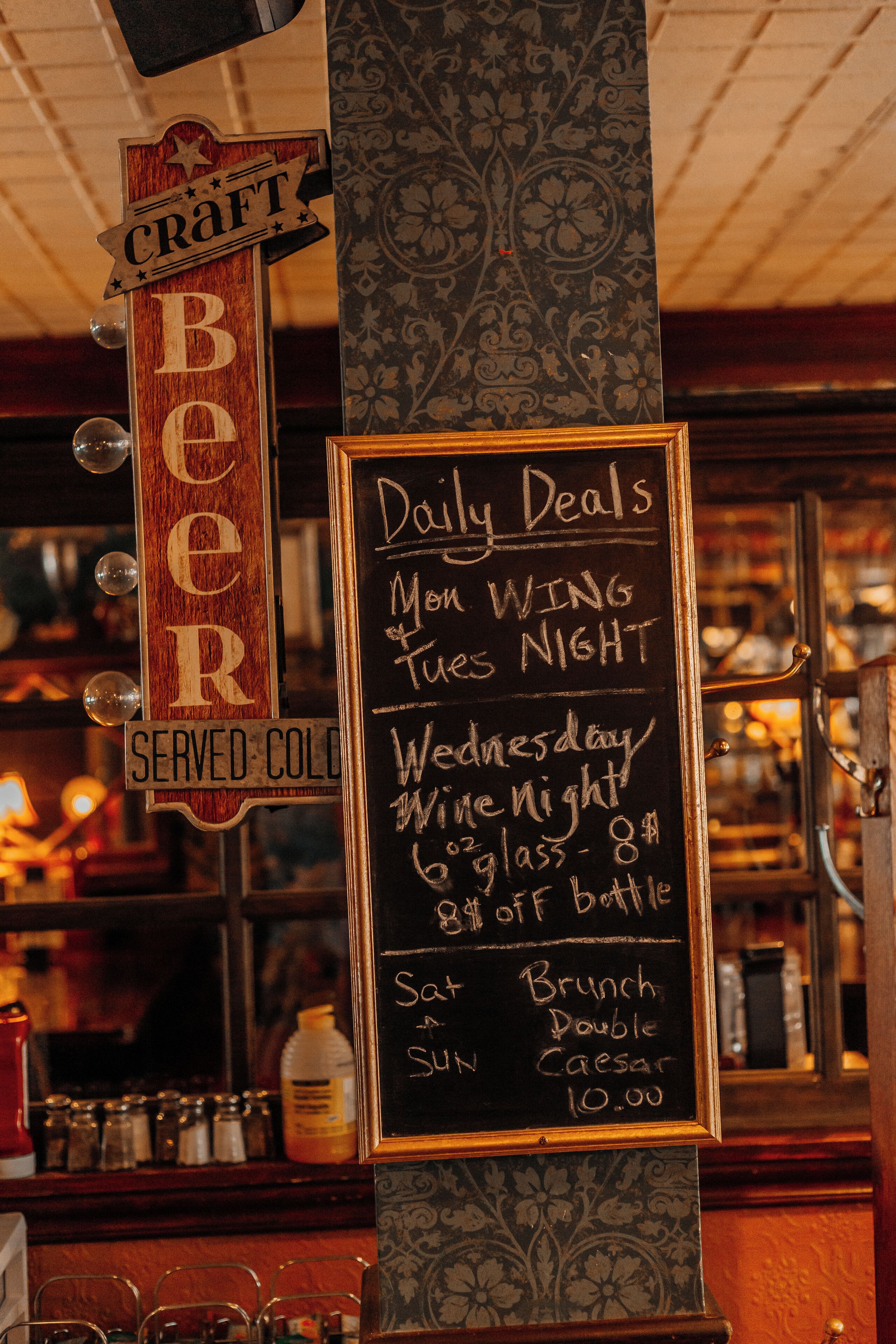Um bar com uma placa de madeira que indica cerveja, uma lousa com promoções diárias e um pilar decorativo.
