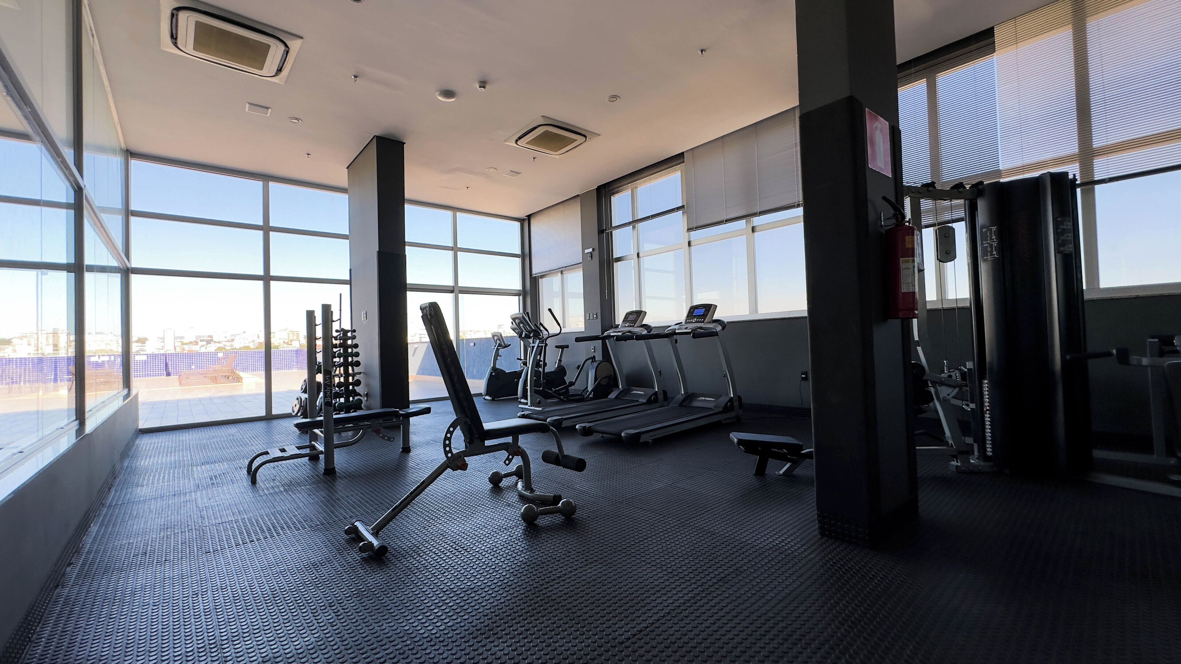 Academia com esteiras, máquinas de musculação e vista da cidade por grandes janelas.
