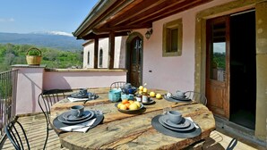 Mesa de madeira posta para o café da manhã, com pratos, xícaras e uma tigela de frutas em uma varanda com vista para uma paisagem montanhosa.