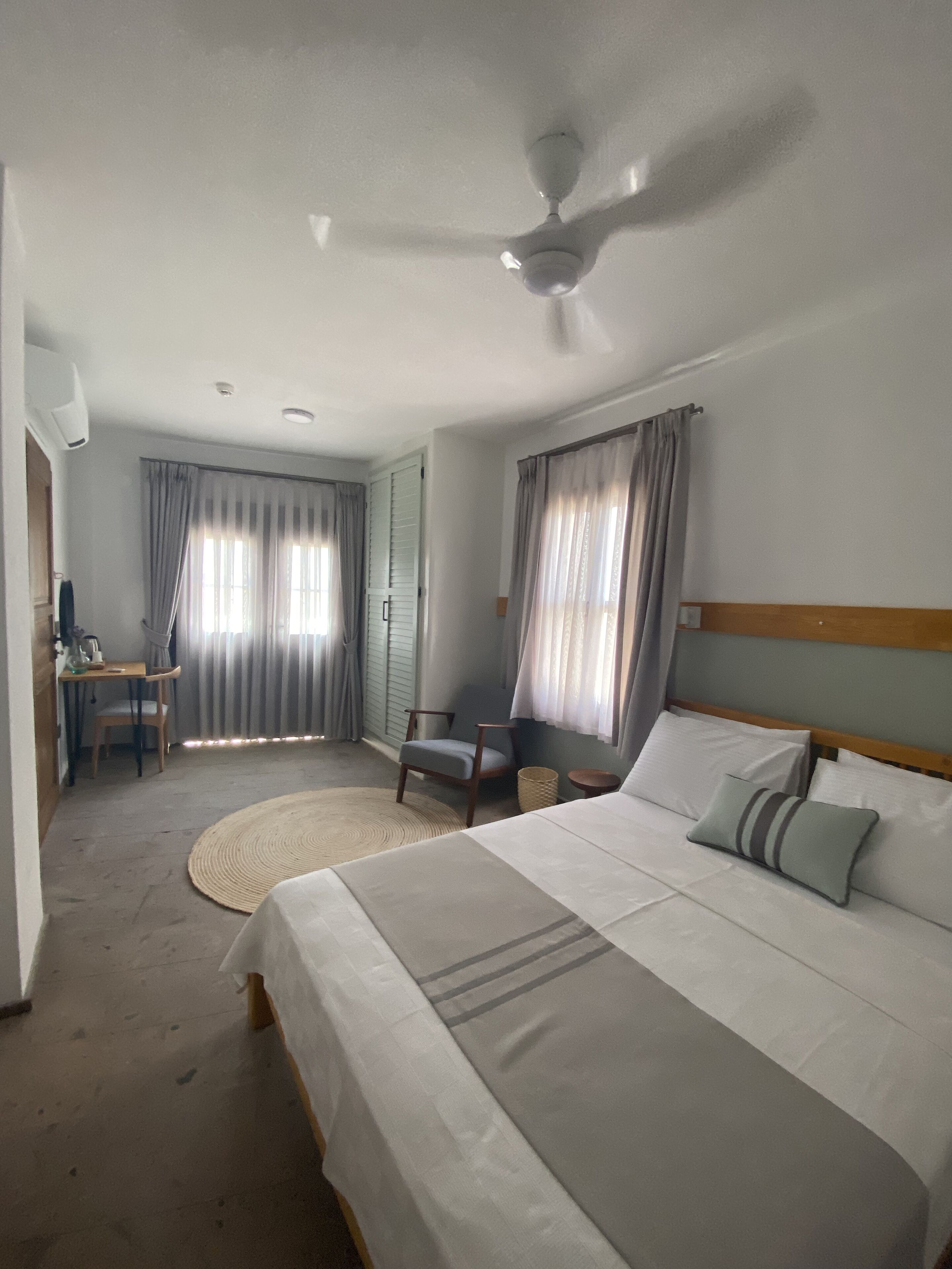Quarto de hotel com uma cama grande, uma escrivaninha, uma cadeira e um ventilador de teto.