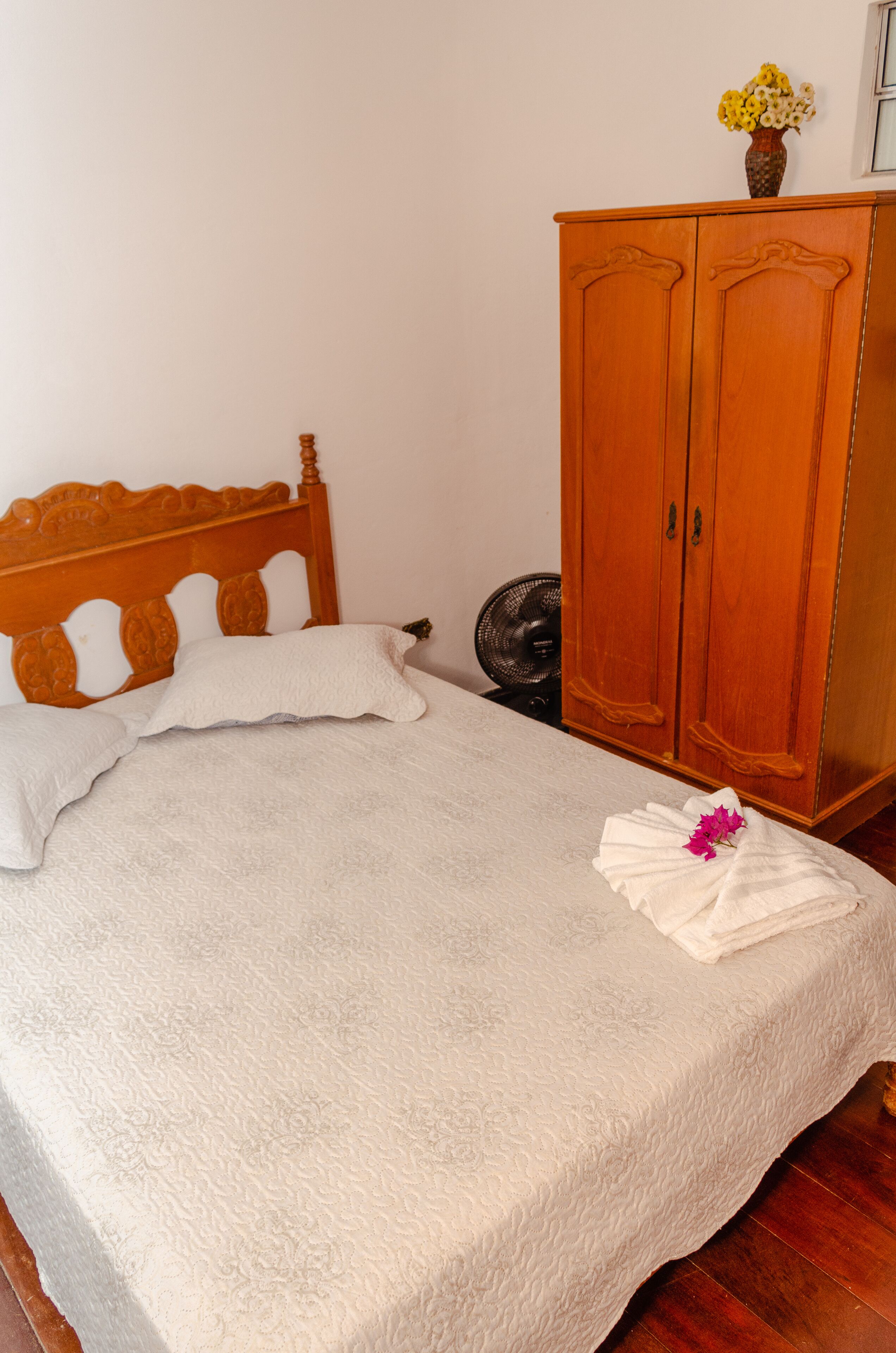 Cama bem arrumada com colcha e travesseiros brancos, um guarda-roupa de madeira e um pequeno vaso com flores em cima.