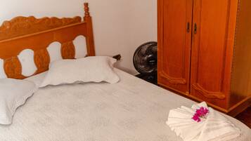 Cama bem arrumada com colcha e travesseiros brancos, um guarda-roupa de madeira e um pequeno vaso com flores em cima.
