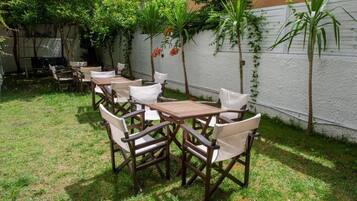 Um pátio externo com mesa de madeira e cadeiras brancas, rodeado por vegetação e uma parede branca.