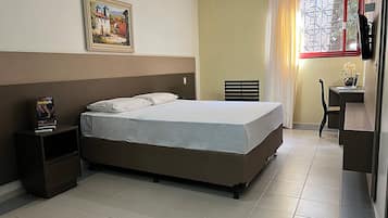 Quarto de hotel com uma cama, uma escrivaninha com um laptop, uma cadeira, um quadro na parede e um ar condicionado.