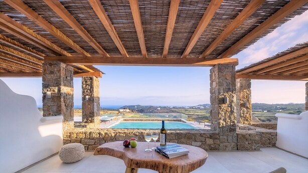 Uma mesa de madeira com um livro, dois copos e frutas, numa área ao ar livre e ensolarada, com colunas de pedra e vista para uma piscina e paisagem.