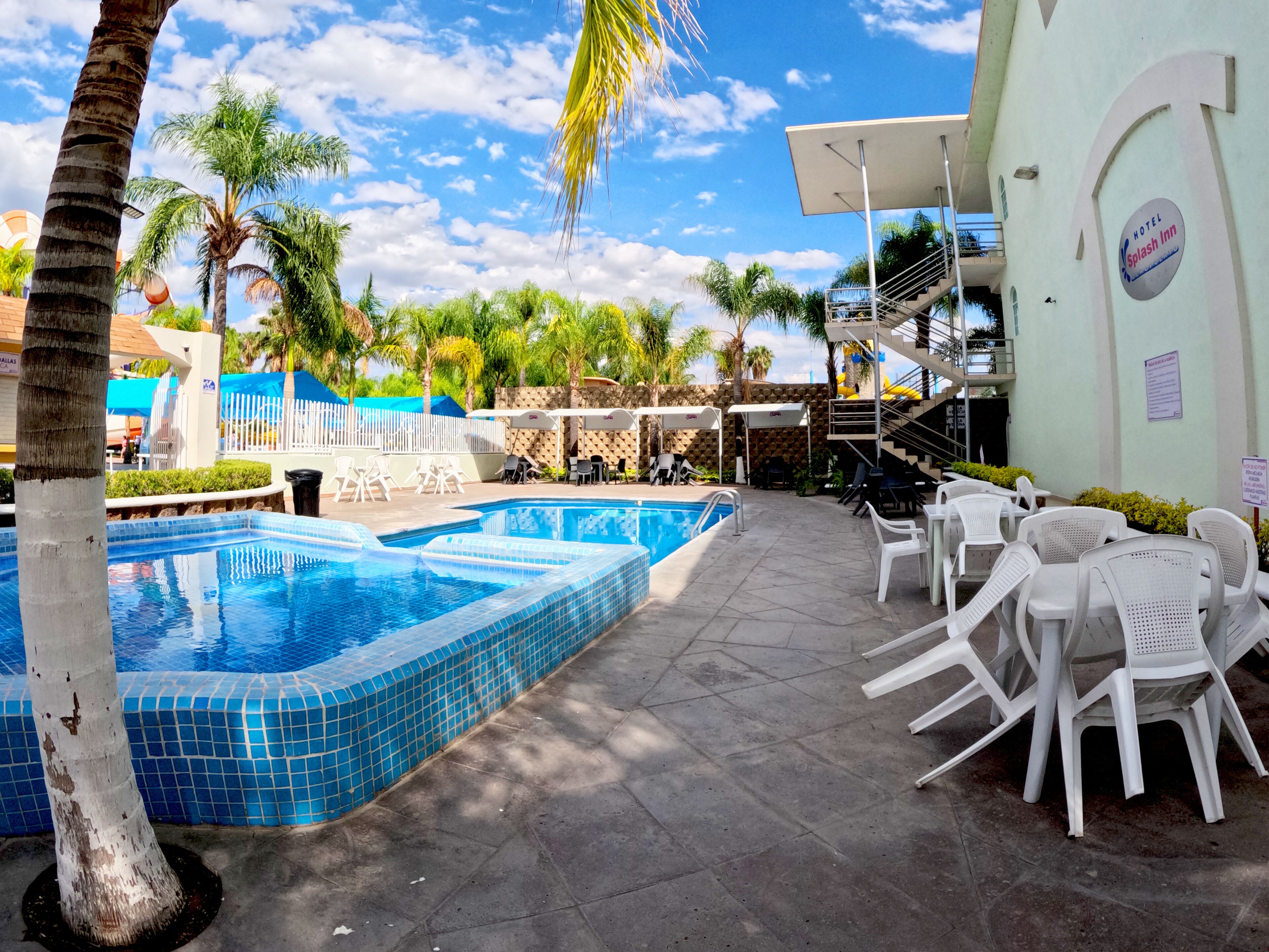 Piscina com bordas azulejadas, cercada por palmeiras, cadeiras de plástico branco e uma área de estar coberta.