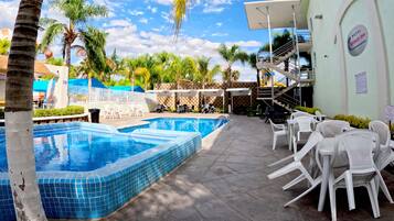Piscina com bordas azulejadas, cercada por palmeiras, cadeiras de plástico branco e uma área de estar coberta.