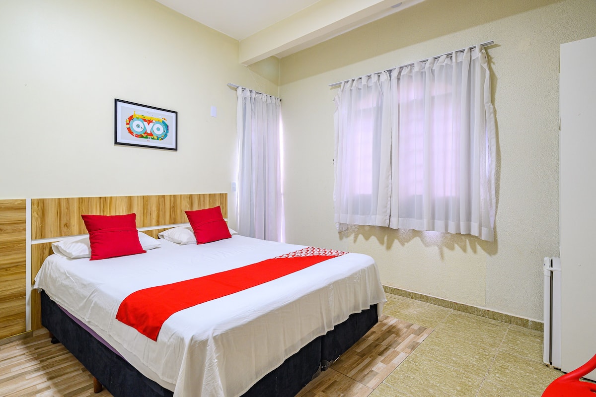 Quarto de hotel com cama, almofadas vermelhas, colcha branca, quadro pendurado na parede e cortinas brancas nas janelas.